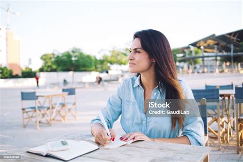 캐주얼 한 옷을 입고 아름다운 사려 깊은 여성 기사를 만들기위한 카피 북에서 메모를 만드는 젊은 30 대 여성은 카페 테라스에서 야외에서 앉아 작업을 설계하기위한 스케치를