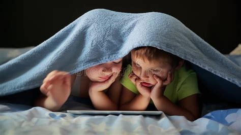 Little Boy And Girl Play On The Tablet While Lying Under A Blanket
