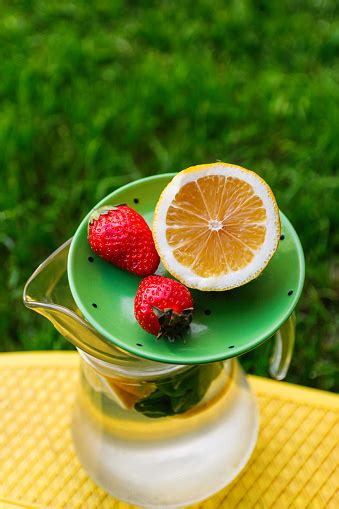 레몬과 두 개의 딸기가 레모네이드 유리 용기에 서 있는 데포커스 접시에 슬라이스 레몬과 민트 잎을 옐로우 보드에 얹습니다 흐린