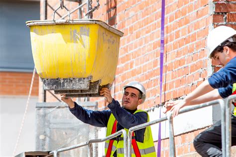 보호 및 안전 장비를 착용한 작업자가 높이에서 크레인 체인에 콘크리트 혼합물이 있는 컨테이너를 들어 올리도록 지시합니다 2명에 대한 스톡 사진 및 기타 이미지 Istock