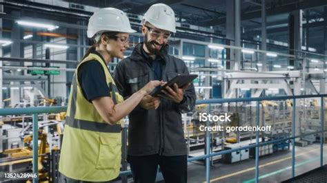 태블릿 컴퓨터를 사용하여 높은 가시성 조끼에 남성 전문가 및 여성 자동차 공장 엔지니어 자동차 산업 제조 시설은 자동차 생산에 종사하고 있습니다 조립 공장의 다양성 제조에