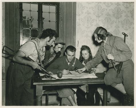Three Male And Two Female Italian Partisans Seated At A Table Inside A Building Looking Over A