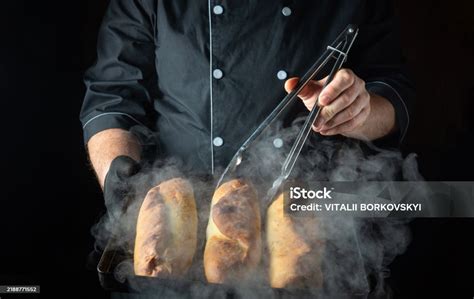 증기가 가득한 따뜻한 주방에서 셰프는 집게를 사용하여 베이킹 트레이에서 황금빛 갈색 빵을 들어 올리며 빵 만드는 기술과 요리 전문 기술을 보여줍니다 빵에 대한 스톡 사진 및