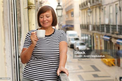 중년 여성은 아침에 유럽 도시 거리 전망이 있는 발코니에 서서 커피나 차 한 잔을 들고 있습니다 리조트에서 휴가를 즐기는 성숙한