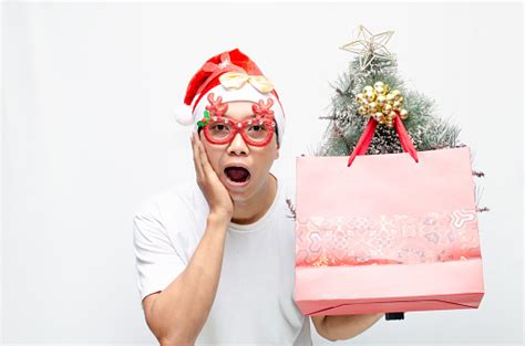 산타 클로스 모자와 크리스마스 안경을 쓴 기쁨에 찬 젊은 아시아 남자가 크리스마스 트리와 함께 빨간 쇼핑 가방을 들고 있습니다 12월에 대한 스톡 사진 및 기타 이미지