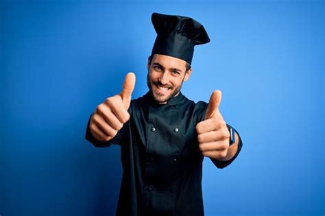 수염을 입은 젊은 잘생긴 요리사 남자는 손으로 긍정적 인 제스처를하고 승인 파란색 배경위에 밥솥 유니폼과 모자를 입고 엄지 손가락으로 미소를 짓고 성공을 위해 행복합니다