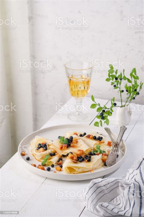 접시에 블루 베리 복숭아 민트를 곁들인 팬케이크 흰색 나무 테이블입니다 밝은 배경입니다 노란 꽃과 노란 음료가있는 소박한 테이블 제공 Maslenitsa에 대한 스톡 사진