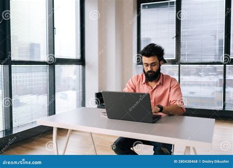 Concentrated Indian Programmer Male In Eyeglasses Working On Laptop Sitting At Desk In Light