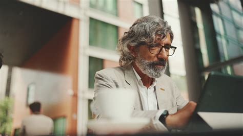 회색 재킷을 입고 안경에 수염을 가진 성숙한 사업가 태블릿에서 일하고 야외 카페에 앉아있다 자연의 카페 테이블에 앉아있는