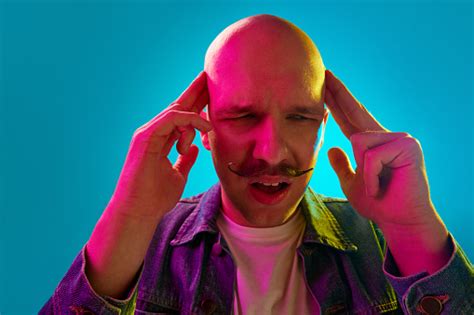 콧수염을 기른 젊은 대머리 남자의 초상화는 네온 불빛의 파란색 스튜디오 배경에 사려 깊은 집중된 얼굴로 서 있다 20 24세에 대한 스톡 사진 및 기타 이미지 Istock