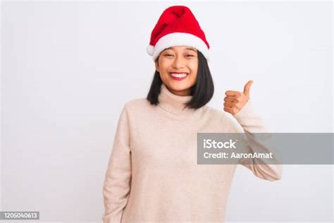 고립 된 흰색 배경 위에 크리스마스 산타 모자를 쓰고 행복한 엄지 손가락으로 손을 들고 제스처를 하는 젊은 아름다운 중국 여성 성공을 보여주는 카메라를 보고 표정을