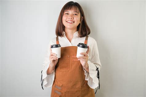 긴 흰색 소매 셔츠와 갈색 앞치마를 입은 흥분한 젊은 아시아 여성이 웃는 얼굴 표정으로 커피 음료를 들고 고객에게 제공하고 있습니다 흰색 배경 위에 고립되어 있습니다 25
