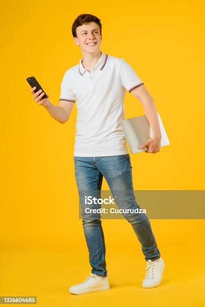 흰색 티셔츠를 입은 젊은 행복한 남자의 초상화가 노란 배경에 노트북 컴퓨터와 스마트폰을 들고 미소짓고 있습니다 20 29세에