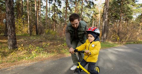 젊은 아버지는 안전 헬멧과 캐주얼웨어를 입은 어린 아들이 공원의 아스팔트 도로를 따라 균형 잡힌 자전거를 타는 것을 도우면서 약간 구부렸다 사진 Unsplash의 운동 이미지
