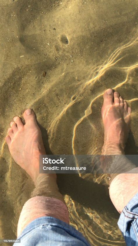 바다에서 노를 젓는 흰색 영국 남자의 발과 롤업 블루 데님 청바지의 이미지는 황금 해변 모래 파도랩핑 해안 해안선에 젖은 얕은 바다 물을 받고 맨발로 발가락 잔물결 반사