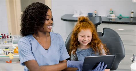 의학 소아 치과 및 구강 관리 개념 현대 치과 진료소에서 행복한 아이 환자 곱슬 빨간 머리 여고생에게 태블릿 Pc 컴퓨터를 보여주는 여성 웃는 아프리카 치과 의사