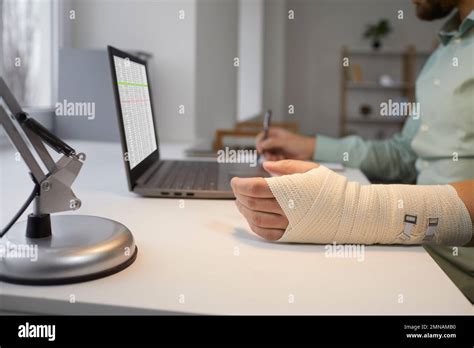 Bearded Man With Bandaged Broken Arm Is Working At Laptop Sitting At His Desk And Making Notes