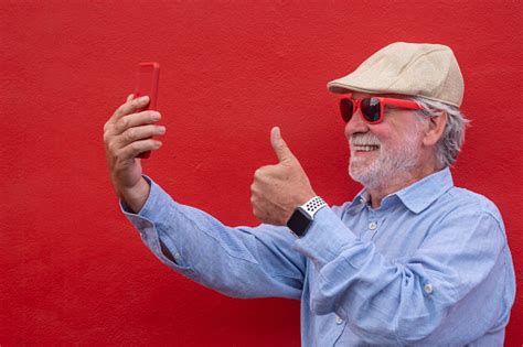 수염과 모자를 가진 수석 흰색 머리 남자는 빨간색 배경에 서 셀카에 대한 자신의 스마트 폰을 사용하여 미소 남자에 대한 스톡 사진 및 기타 이미지 Istock