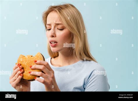 Unhappy Beautiful Blonde Girl Eating Hamburger On Camera Isolated Over Blue Background Stock
