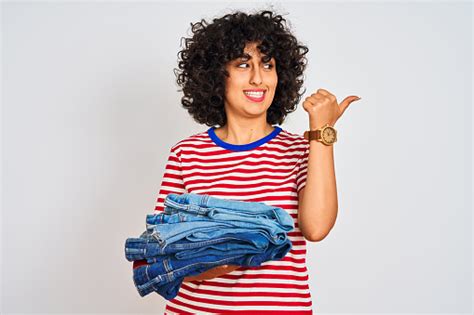 젊은 아랍 상점 주인 여자 와 곱슬 머리 들고 청바지 위에 고립 된 흰색 배경 가리키는 고 행복 한 얼굴 미소와 측면에 엄지 손가락으로 표시 더미에 대한 스톡 사진 및 기타