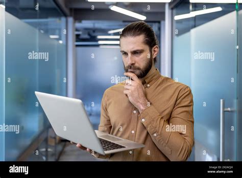 A Thoughtful Male Programmer Holding A Laptop And Analyzing Data In A Contemporary Office