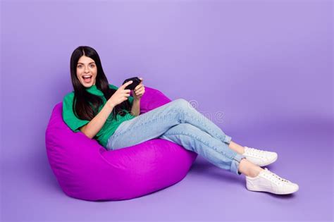 Joyful Young Woman Enjoying Leisure Time On A Purple Bean Bag Holding Game Controller In A Joyful Young Woman Enjoying Leisure Time On A Purple Bean Bag Holding Game Controller In A