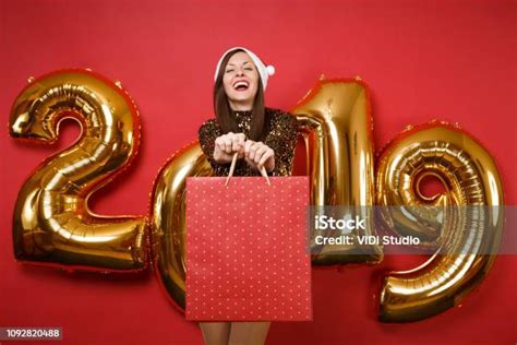 반짝 반짝 드레스에서 메리 산타 여자 크리스마스 모자 밝은 빨간 벽 배경 황금 번호 공기 풍선 스튜디오 초상화에 고립 된 쇼핑백 개최 행복 한 새 해 2019 휴일 파티