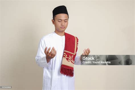 해골 모자를 쓴 흰 셔츠를 입은 매력적인 아시아 무슬림 남자의 초상화가 손을 들고 간절히 기도하고 있다 회색 배경에 격리된