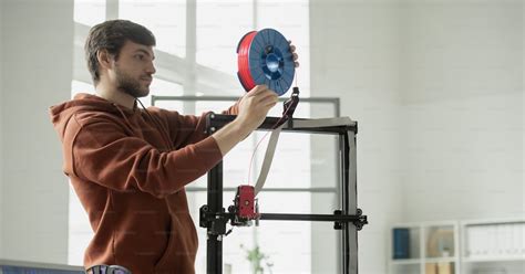 기하학적 도형을 인쇄하는 동안 3d 프린터에 빨간색 필라멘트가 있는 새 스풀을 넣는 청년 사진 Unsplash의 초상 이미지
