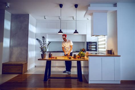 잘 생긴 미소 백인 요리사의 전체 길이 유니폼과 부엌에 서서 양파를 절단 머리에 산타 모자와 함께 부엌 카운터에는 다양한 종류의 채소가 있습니다 부엌에 대한 스톡 사진 및