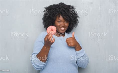 젊은 아프리카계 미국인 여자 회색 그런 지 벽 먹는 동안 핑크 도넛 큰 행복 확인 서명 하 고 미소 손가락 우수한 로그인으로