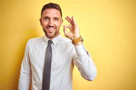 노란색 고립 된 배경 위에 우아한 흰색 셔츠를 입고 젊은 잘 생긴 사업가 는 손과 손가락으로 확인 기호를하고 긍정적 인 미소