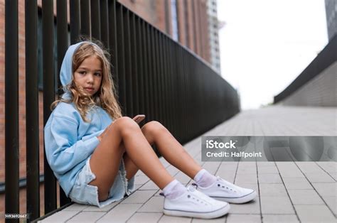 후드를 가진 작은 금발 소녀는 보도에 앉아 스마트 폰을 들고 카메라를보고 거리에 대한 스톡 사진 및 기타 이미지 거리