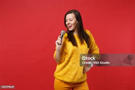멋진 노란색 모피 스웨터에 아주 젊은 여자의 초상화 스튜디오에서 밝은 빨간 벽 배경에 고립 된 마이크에서 노래 사람들이 진심으 감정 라이프 스타일 개념 복사 공간을 비웃는