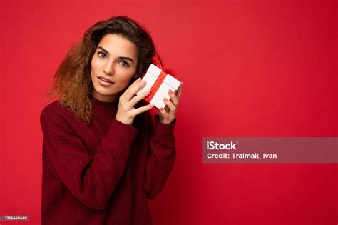 아름다운 사려 깊은 젊은 갈색 머리 곱슬 여성 사람 고립 된 빨간 배경 벽 입고 빨간 스웨터 들고 선물 상자 보고 카메라