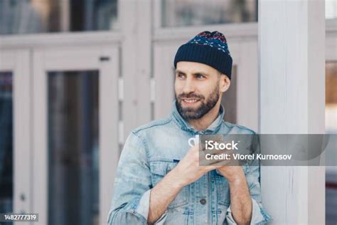 잘 생긴 웃는 남성의 수평 샷은 행복하게 멀리 보인다 모자와 청바지 재킷을 입고 커피 잔을 들고 즐거운 무언가를 통지 집에서 휴식 사람 라이프 스타일 및 레크리에이션 개념