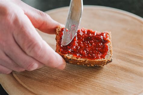 라즈베리 잼을 곁들인 롤빵 손은 곡물 롤빵에 라즈베리 잼을 뿌립니다 아침 식사 준비 라즈베리 잼을 곁들인 통곡물 롤빵 전통적인 아침 식사 음식 달콤한 롤빵에 대한 스톡 사진