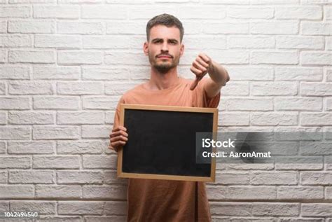 성 난 얼굴 아래로 엄지 싫어 보여주는 음수 부호 거부 개념 칠판을 들고 벽돌 벽을 통해 젊은 성인 남자 개념에 대한 스톡 사진 및 기타 이미지 Istock