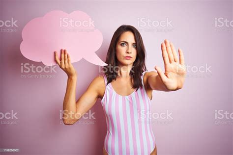 스트라이프 핑크 수영복을 입고 진지하고 자신감있는 표정으로 정지 표지판을하는 열린 손으로 분홍색 배경 위에 말하는 풍선을 들고있는 아름다운 여성 갈색 머리에 대한 스톡 사진