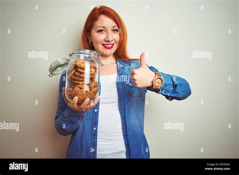 Young Beautiful Redhead Woman Holding Jar Of Cookies Over White