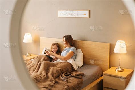 镜子里的倒影是年轻快乐的妈妈和可爱快乐的女儿一起躺在床上读着儿童读物准备睡觉。素材 花瓣网