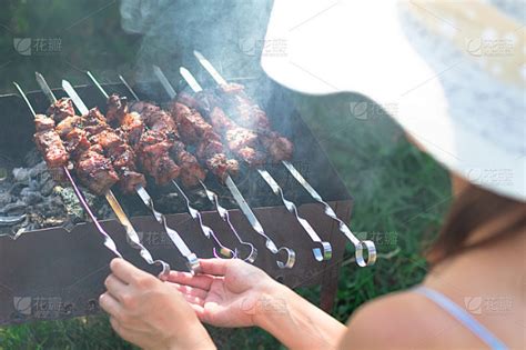一个戴着草帽的女孩在一个阳光明媚的夏日，在绿叶的映衬下，在烤架上准备着芳香的新鲜肉