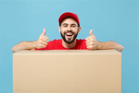 파란색 배경 스튜디오 초상화에 고립 된 빨간색 유니폼을 입은 배달 남자 택배 딜러로 일하는 모자 티셔츠 프린트의 남성 직원은 빈 골판지 상자를 들고 있습니다 서비스 개념