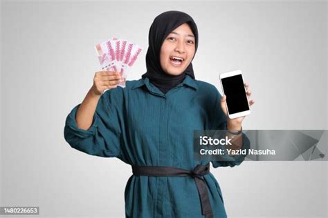 히잡을 쓴 흥분한 아시아 무슬림 여성의 초상화는 빈 화면 모형 휴대폰을 보여주면서 10만 루피아를 보여준다 재정 및 저축 개념 흰색 배경에 격리된 이미지 18 19세에 대한