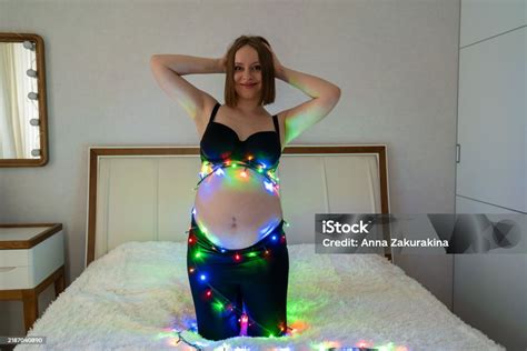 임신 한 배꼽 아기 범프에 조명 화환을 가진 임산부는 집에서 침대에 앉아있다 크리스마스 임신 즐거운 성탄절 보내시고 새해 복 많이 받으세요 겨울철 출산 예정 12월 31일에