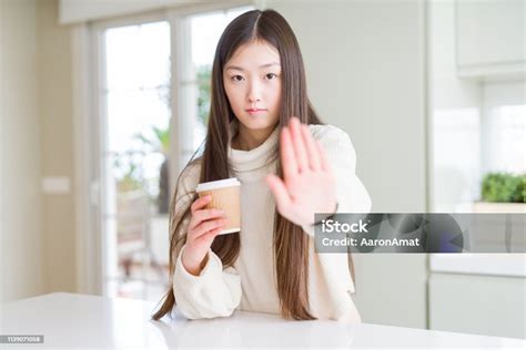 아름 다운 아시아 여자에 커피를 마시는 오픈 손으로 테이크 아웃 종이 컵을 하 고 심각 하 고 자신감 있는 표현 국방 제스처와 함께 중지 표시 가정 생활에 대한 스톡 사진