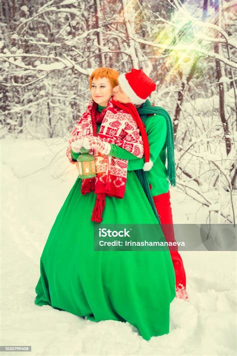 젊은 부부 남자와 여자 남편과 아내는 산타의 도우미 가슴 가득 선물 및 거 대 한 사탕으로 눈 아래 겨울 숲에서의 엘 프의 전형적인 꽃의 의상에서 걷고 있다 2명에 대한