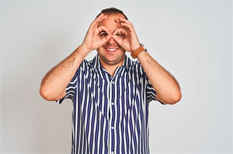 고립된 흰색 배경 위에 서있는 파란색 줄무늬 셔츠를 입은 청년은 혀를 내밀고 손가락을 통해 눈을 내밀고 있는 쌍안경처럼 괜찮은 제스처를 하고 있습니다 미친 표정 긍정적인