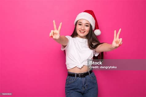 Pnk 배경을 통해 고립 된 승리 기호를하고 카메라에 윙크 행복한 얼굴로 웃는 크리스마스 모자를 입고 젊은 아시아 여성 겨울에 대한 스톡 사진 및 기타 이미지 Istock