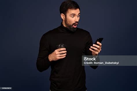 블루 월 배경 스튜디오 초상화에 고립 포즈 트렌디 한 스웨터에 미소 젊은 남자 남자는 종이 커피 컵이나 차를 들고 전화를보고